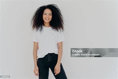 곱슬 곱슬 한 어두운 머리를 가진 미소 짓는 여자의 사진 슬림 한 그림을 가지고 흰색 티셔츠와 검은 청바지를 입고 기쁜 얼굴 표정 흰색 배경에 대한 모델 광고 콘텐츠에 대한