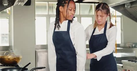 In A Professional Kitchen A Female Chef Assists Her Friend In Tying An