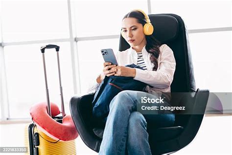 공항 라운지에서 헤드폰을 끼고 여행 가방을 옆에 두고 스마트폰을 확인하고 있는 매력적인 여성 20 24세에 대한 스톡 사진 및 기타 이미지 Istock