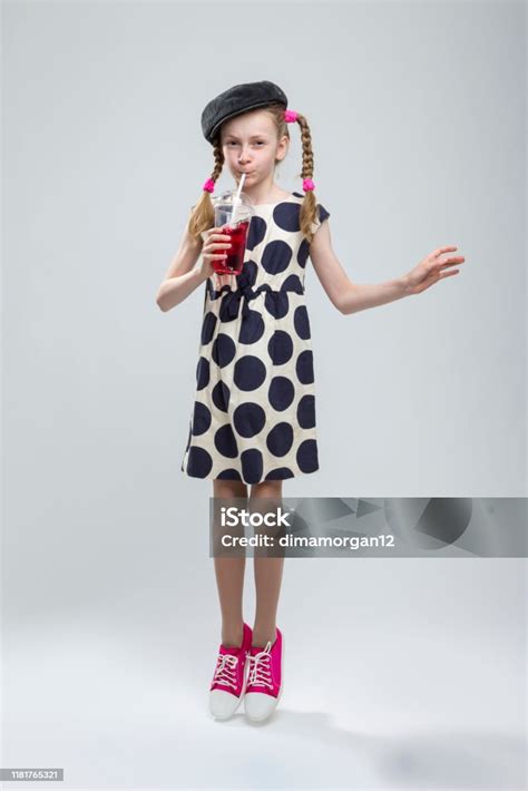 회색 벨벳 모자와 빨간 주스 한 잔과 폴카 도트 드레스에 포즈를 취하는 피그 테일과 백인 소녀점프의 전체 길이 초상화 빨대를