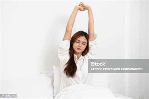 하얀 잠옷을 입은 여자가 주말 아침에 일어나 휴식을 취하고 노트북 휴대폰을 가지고 놀며 하얀 침실 안의 유리에서 빵을 먹고 차를 마신다 아침 휴가 개념입니다 갈색 머리에