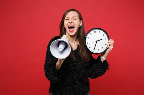 검은 모피 스웨터에 젊은 여자를 웃 고 스튜디오에서 밝은 빨간색 벽 배경에 고립 된 라운드 시계를 들고 확성기에 비명 사람들은 진심으로 감정 라이프 스타일 개념입니다 복사