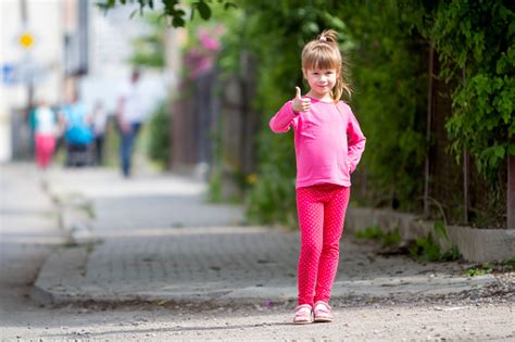 분홍색 캐주얼 의류와 긴 금발 조랑말 꼬리에 귀여운 작은 소녀의 전체 길이 초상화 흐리게 밝은 햇볕이 잘 드는 거리 배경에 엄지