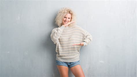 그런 지에 곱슬 머리 젊은 금발의 여 자가 회색 배경 손으로 크고 큰 크기 표시 측정 기호를 보여주는 몸짓 카메라를 보고 웃 고