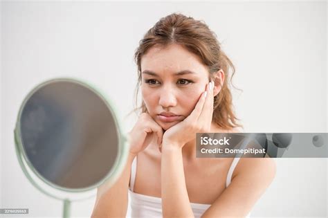 젊고 아름다운 아시아 여성이 거울에 비친 얼굴 피부를 확인하고 얼굴에 주근깨 주름 여드름 흉터에 대해 걱정하고 있습니다 레이디 뷰티 셀프 케어 스킨 케어 문제 솔루션 제품