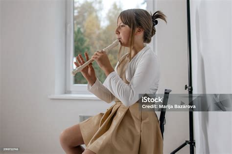 소녀는 밝은 방에서 플루트를 연습하고 흰색 배경에 검은 의자에 앉아 연주합니다 어린이 음악가 개념입니다 학교 밖 활동 가정 원격 학습 상의 View 8 9 살에 대한 스톡