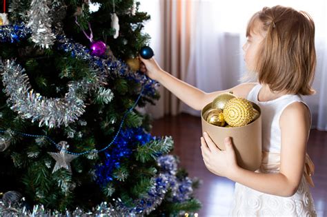 흰색 드레스를 입은 어린 소녀는 금색과 파란색 크리스마스 볼로 크리스마스 트리를 장식하고 커튼이있는 방에서 집에서 블링합니다 겨울 방학에 가족 활동 황소의 새해 사진 촬영