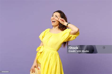 노란 드레스를 들고 있는 쾌활 한 젊은 여자 여름 모자를 보여주는 승리 기호 옆에 고립 된 파스텔 바이올렛 벽 배경 사람들은 진심으로 감정 라이프 스타일 개념입니다 복사