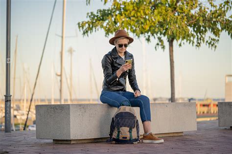 고급 모자 선글라스 가죽 재킷에 짧은 머리를 가진 30대 금발 여성이 바르셀로나의 요트 마린 옆 벤치에서 스마트폰을 사용하고 있다 30 39세에 대한 스톡 사진 및 기타