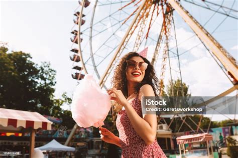 선글라스와 생일 모자에 어두운 곱슬 머리를 가진 미소 짓는 여성은 면 사탕을 손에 들고 서서 행복하게 옆으로 바라보며 관람차가있는 놀이 공원에서 시간을 보내고 있습니다