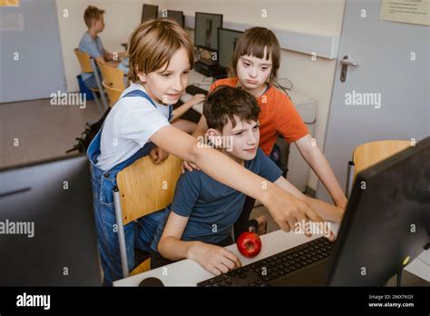 Curious Girl Pointing At Computer Near Boy And Female Friend With Disability In Classroom Stock