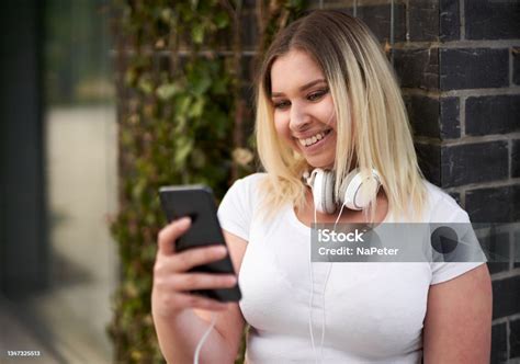 행복한 젊은 쾌활한 밀레니엄 학생 소녀는 검은 벽돌 벽 앞에 서서 그녀의 목에 헤드폰으로 스마트 폰에서 채팅 십대와 사람들의 라이프 스타일과 기술 개념 20 24세에 대한