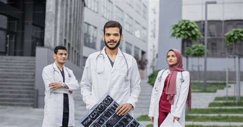 다인종 3명의 의사로 구성된 자신감 넘치는 팀이 스크럽과 코트를 입고 야외에 서 있습니다 남성 아랍 의사 지도자는 단층 촬영 스캔을 손에 들고 미소로 카메라를 바라보고