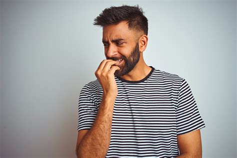 검은 줄무늬 티셔츠를 입은 젊은 인도 남자가 고립 된 흰색 배경 위에 서서 손톱을 물고 손에 손을 대고 스트레스를 받고 긴장합니다 불안 문제 T 셔츠에 대한 스톡 사진 및