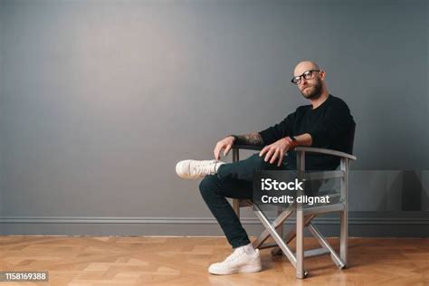 스튜디오의 회색 벽 배경에 자신감있는 포즈를 취하고 의자에 다리 위에 다리를 대고 앉아있는 현대인 검은색에 대한 스톡 사진 및