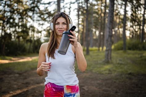 한 여성 행복한 백인 여성 숲이나 숲에 공원에 서서 자연 훈련을하는 동안 보충 쉐이커와 헤드폰으로 브레이크 워터 음료와 수분 공급 실제 사람들은 공간을 복사합니다 개념에