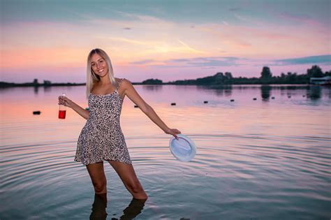 그녀의 손에 모자를 가진 젊은 미소 여자는 아름다운 일몰에 물 위를 걷는 것을 즐긴다 금발 머리에 대한 스톡 사진 및 기타 이미지 금발 머리 드레스 마시기 Istock