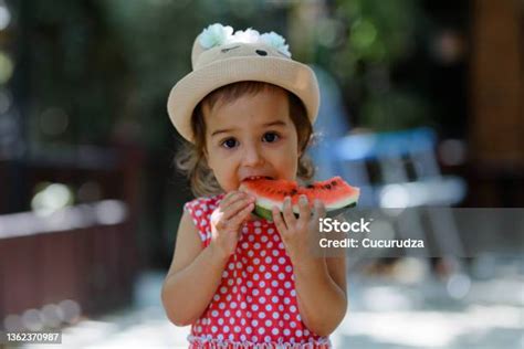 흰색 물방울 무늬와 모자가있는 빨간 드레스를 입은 어린 2 살 소녀는 수박의 매우 재미있는 조각을 먹고 있습니다 개념에 대한 스톡 사진 및 기타 이미지 Istock