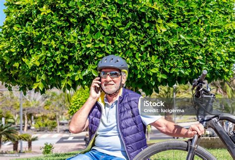 휴대 전화를 사용하여 공원에서 야외에 앉아있는 전기 자전거를 가진 노인의 초상화 자전거 가까이에서 헬멧을 쓰고 핸드폰으로 이야기하는 노인 할아버지 미소 짓기 70 79세에