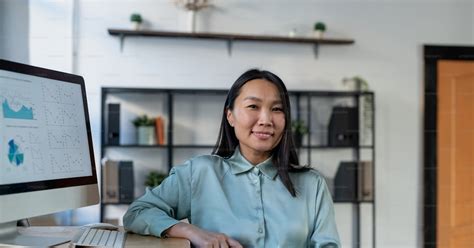 스마트 캐주얼웨어를 입은 젊은 아시아 여성 경제학자가 컴퓨터 모니터와 재무 데이터를 화면에 표시하며 직장에 서 있다 사진