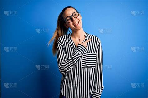美丽的金发女人，蓝色的眼睛，穿着条纹衬衫和眼镜在蓝色的背景愉快地与微笑的脸指向与手和手指向上的一边与