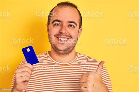확인 기호를하고 큰 미소행복 고립 된 노란색 배경 위에 서 파란색 신용 카드를 들고 젊은 남자 손가락으로 엄지 손가락 우수한 기호 고객에 대한 스톡 사진 및 기타 이미지