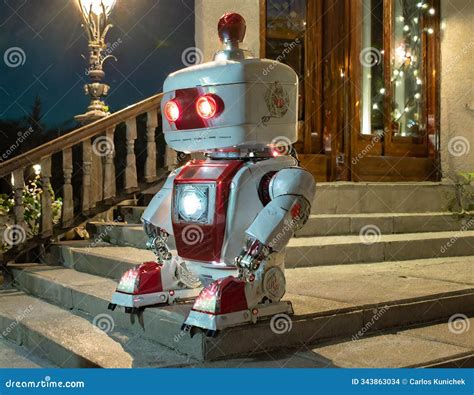 A Small Retro Futuristic Robot With A Cute Chubby Design Sitting On The Steps Of A Warmly Lit