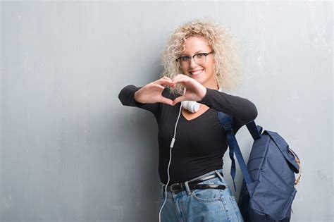 젊은 금발 여자 배낭과 심장 기호와 손으로 모양을 보여주는 사랑에 웃 고 헤드폰 입고 그런 지 회색 벽에 낭만적인 개념입니다 가방에 대한 스톡 사진 및 기타 이미지 Istock