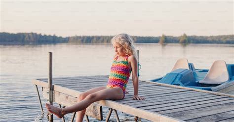 재미있고 행복한 귀여운 백인 금발 소녀가 호수 옆 나무 갑판 부두에 앉아 있다 수영복을 입고 웃고 있는 아이가 물에 다리를 담그고 튀고 있다 여름의 재미있는 야외 활동