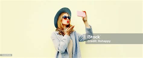 스마트 폰으로 셀카를 찍고 입술을 불고있는 아름다운 행복한 여성의 초상화는 둥근 모자 흰색 배경에 코트 재킷을 입고 공기 키스를 보냅니다 20 29세에 대한 스톡 사진 및