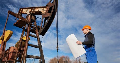 석유 펌프 잭에서 유전 계획을 들고 있는 석유 노동자의 측면 모습 작업복을 입고 헬멧을 쓴 드릴 작업자가 오일 펌핑 장치 근처에 서 있습니다 석유 산업과 기름 적출의