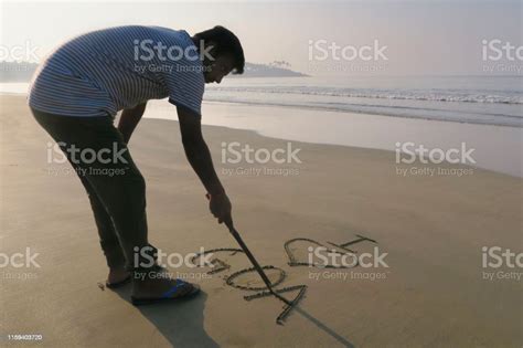 스트라이프 티셔츠와 올리브 그린 바지를 입고 햇볕이 잘 드는 해변에 쓰는 인도 남자의 이미지 와 함께 바다 팔로렘 비치 고아 인도 나무 막대기로 황금 모래에 필기의 개념