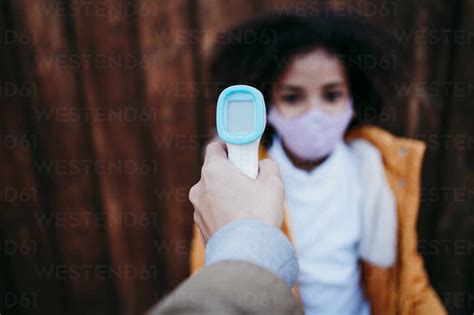 Woman Using Infrared Thermometer While Checking Girl Temperature Against Wooden Wall Stock Photo