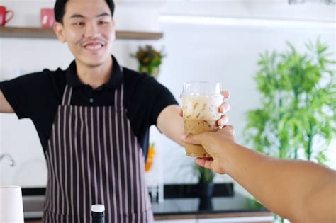 커피숍카운터바에서 아이스 라떼 커피 한 잔을 마시며 미소짓는 아시아 바리스타 청년 투명 플라스틱 유리를 들고 손에 초점을