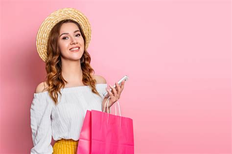 밀짚 모자와 분홍색 배경에 웃고있는 휴대 전화와 쇼핑백이있는 흰색 여름 드레스를 입은 젊은 여성 텍스트를위한 복사 공간 웹 배너 소녀는 온라인 쇼핑 스튜디오에서 사진을