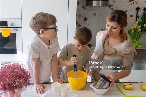 부엌에서 함께 빵을 굽고 있는 엄마와 아이들 어머니와 두 아들이 밝은 부엌에서 노란색 그릇에 재료를 섞고 밀가루를 체로 쳐서 함께 레시피를 만듭니다 3 명에 대한 스톡 사진