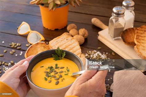 숟가락을 들고 있는 여자 손은 빵 크루통과 호박 씨앗과 함께 제공되는 호박 수프 그릇에 담겨 있습니다 나무 배경 숟가락에 대한 스톡 사진 및 기타 이미지 Istock