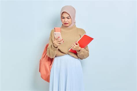 갈색 스웨터를 배낭으로 씌운 젊은 아시아 무슬림 여성 학생에게 충격을 주었고 하얀 배경에 고립 된 책과 휴대 전화를 들고 있었다 학교 개념으로 돌아가기 20 24세에 대한