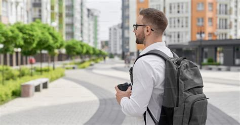 어깨에 배낭을 메고 커피 컵을 들고 서 있는 젊은 수염 난 남자 흰 셔츠를 입고 다층 주택을 바라보는 안경을 쓴 잘생긴 남자 사진 Unsplash의 도시 이미지