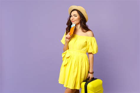 노란 드레스에 웃는 젊은 여자 여름 모자 개최 가방 여권 탑승권 티켓 파스텔 바이올렛 벽 배경에 고립 사람들은 진심으로 감정 라이프 스타일 개념입니다 복사 공간을 모의