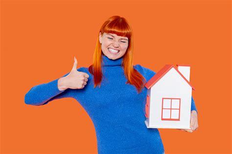 주황색 머리를 한 밝게 차려입은 여성이 주황색 배경에 미니어처 집을 들고 엄지손가락을 치켜세우며 주택 소유나 부동산 성취를