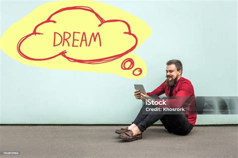 바닥에 앉아있는 빨간 셔츠에 행복 한 수염 사업가 들고 그의 디지털 태블릿을 보고 꿈 꿈 텍스트와 벽에 노란색 구름 모양 건물의 층에 대한 스톡 사진 및 기타 이미지