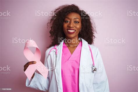 행복한 얼굴 서서 치아를 보여주는 자신감 미소와 함께 미소와 고립 된 분홍색 배경 위에 암 리본을 들고 젊은 아프리카 계 미국인 의사 여자 건강관리와 의술에 대한 스톡 사진