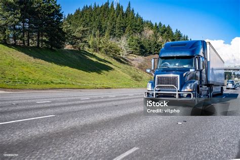 다른 트랙 앞의 평평한 직선 고속도로 도로에서 달리는 드라이 밴 세미 트레일러에서 상업용화물을 운반하는 파이프 그릴 가드가있는 파란색 큰 장비 세미 트럭 미국에 대한 스톡