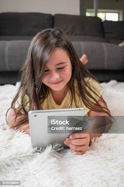 귀여운 어린 소녀는 디지털 태블릿에서 만화를 봅니다 아이는 전자 장치를 사용하여 웃으며 바닥에 누워 있습니다 어린이를위한 실내 레저 고품질 사진 가정 생활에 대한 스톡 사진