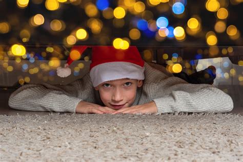 산타 모자를 쓴 예쁜 십 대 소년이 화환 조명으로 침대 아래 바닥에 누워 산타를 초조하게 기다리고 있다 12월에 대한 스톡 사진 및 기타 이미지 Istock