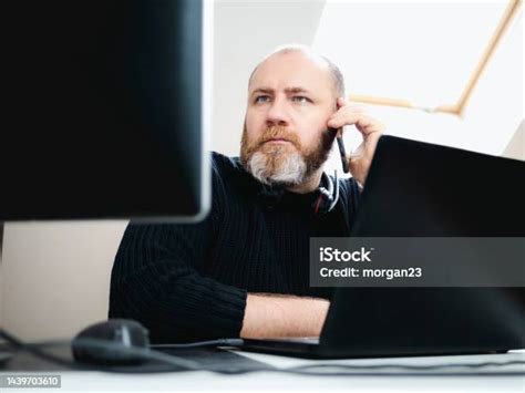 성인 중심의 남성은 노트북에서 원격 작업을 하고 집에서 두 대의 모니터를 사용하는 동안 스마트폰으로 전화를 겁니다 가정 생활에