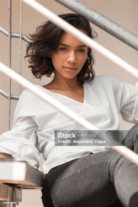 곱슬 머리와 짧은 머리를 가진 젊은 라틴 어 갈색 머리 여자 편안한 손에 들고 흰색 블라우스와 검은 바지를 입고 라틴 문자에