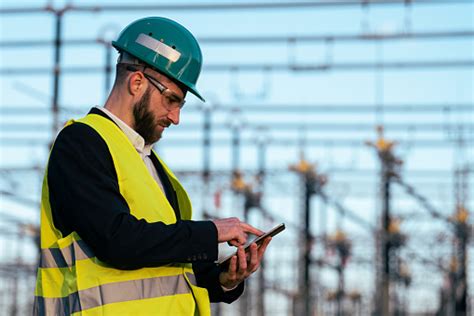 헬멧 정장과 안전 조끼를 입은 전기 엔지니어가 태블릿에서 변전소를 배경으로 데이터를 확인합니다 30 34세에 대한 스톡 사진 및