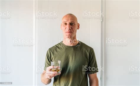 집에서 흰색 벽 앞에 서있는 브랜디 또는 위스키 알코올 음료 한 잔을 들고 녹색 티셔츠를 입고 짧은 머리를 가진 젊은 백인 남자의 초상화 T 셔츠에 대한 스톡 사진 및 기타